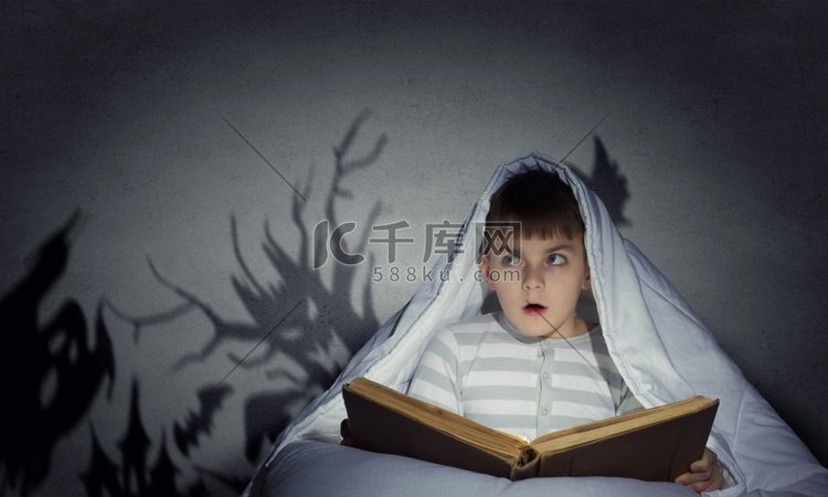 孩子们的噩梦。可爱的小男孩坐在床上，毯子里拿着书高清摄影大图-千库网