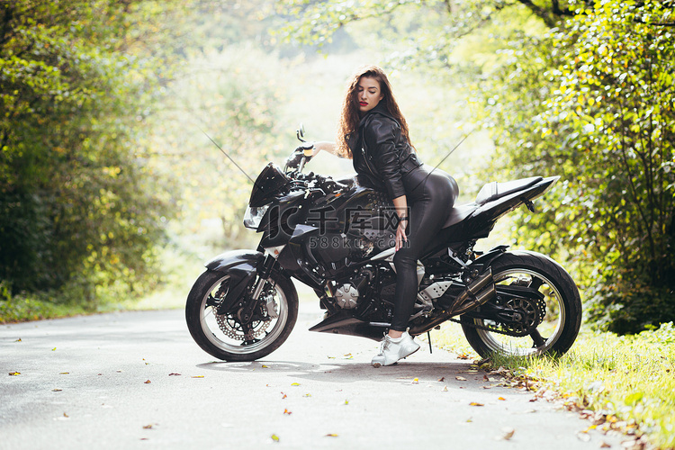 一位美丽的年轻女子坐在摩托车上的肖像，穿着黑色皮衣在大自然、街道、森林里骑着摩托车，在阳光明媚的天气里，充分成长高清摄影大图-千库网