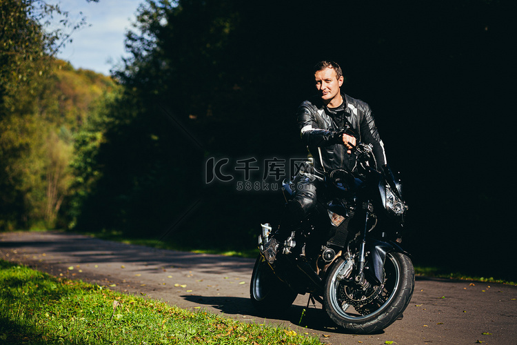 帅气的年轻人坐着骑着黑色皮衣的摩托车，看着镜头，在奔跑中，在路上高清摄影大图-千库网