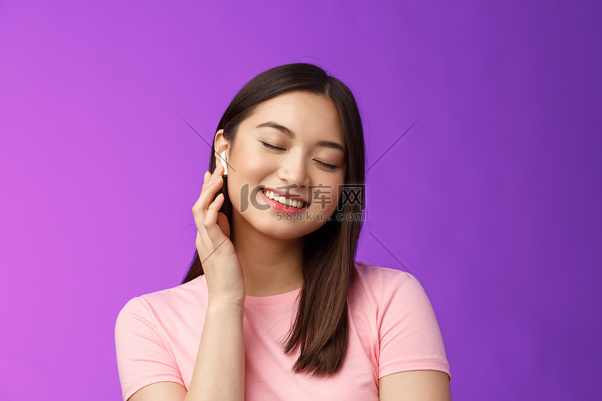 享受听音乐的可爱亚洲女孩,戴上无线耳机,倾斜头闭上眼睛梦幻般的微笑满足,完美的声音,让音量耳机更响亮,站立紫色背景高清摄影大图 千库网
