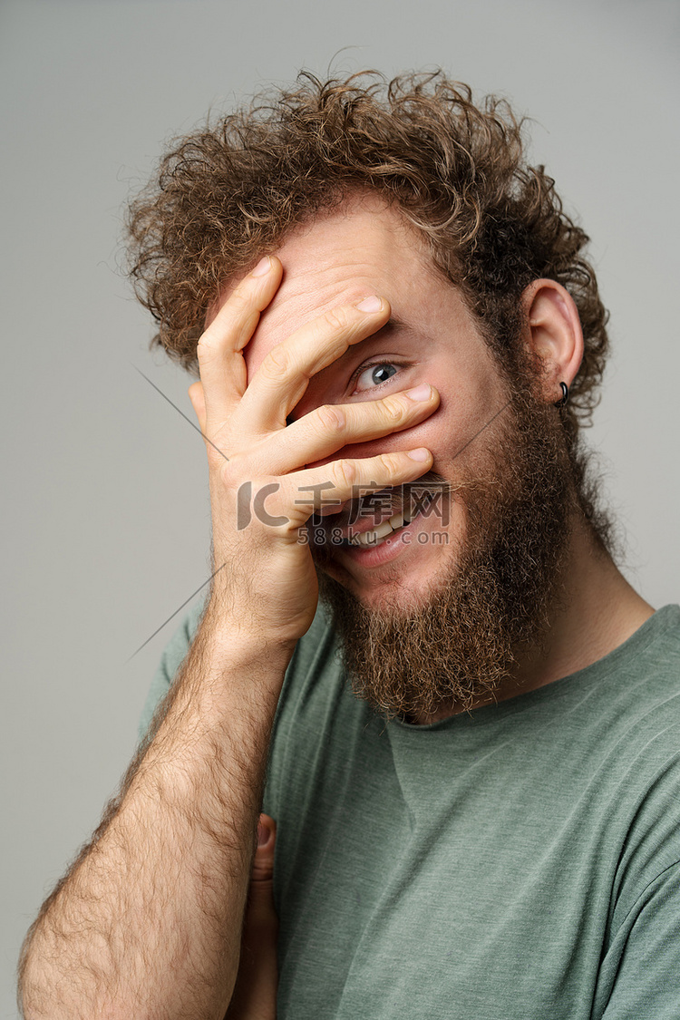 害羞的英俊年轻人用手撬开他的脸，用眼睛撬开他的脸，卷曲的头发穿着橄榄色 T 恤，隔离在白色背景上。高清摄影大图-千库网