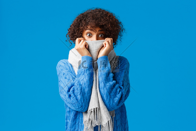 傻乎乎、害怕、脸红的可爱非洲裔美国女人，留着非洲式发型，把脸藏在围巾后面，瞪着眼睛，惊讶或有点焦虑，穿着冬季毛衣站在蓝色背景高清摄影大图-千库网