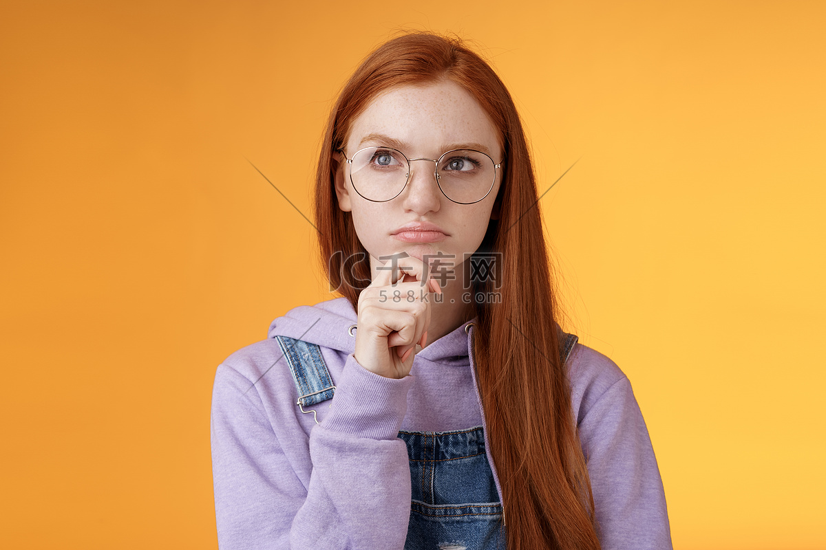 困惑的严肃聪明的年轻红发女孩困惑地思考深焦点问题触摸下巴皱眉看左上角深思熟虑，思考可疑情况，橙色背景高清摄影大图-千库网