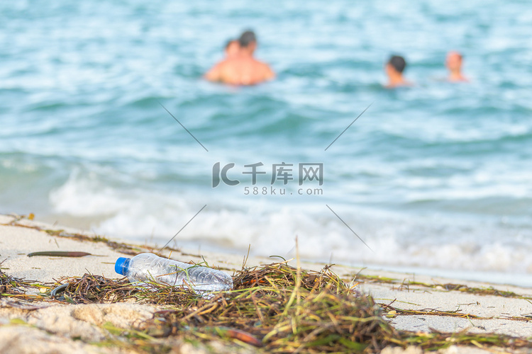 脏沙滩上空塑料水瓶的特写图像，脏沙滩上满是海藻、垃圾和废物，背景是大海中的人高清摄影大图-千库网