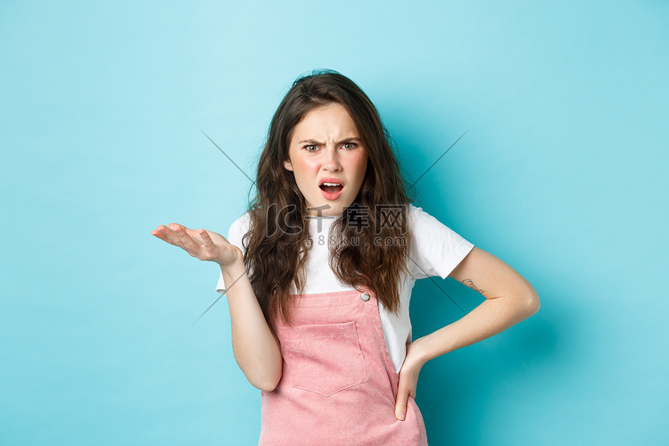 困惑和恼怒的年轻女性举起手，指着一些可怕的东西，站在蓝色背景上，用愤怒的脸困惑地盯着相机的形象高清摄影大图-千库网