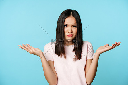 困惑而恼怒的亚洲女孩耸肩，皱眉，张开双手，困惑，无法理解，蓝色背景高清摄影大图-千库网