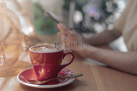 特写热卡布奇诺、拿铁咖啡，手上拿着手机、智能手机的女人模糊地坐在咖啡馆里。高清摄影大图-千库网
