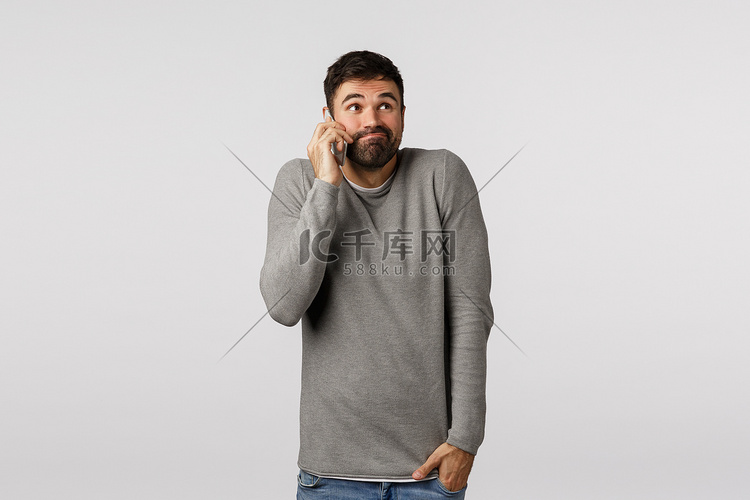 穿着灰色毛衣、优柔寡断、不确定的留着胡须的可爱酷儿家伙，傻笑不确定，答案困惑，手插在口袋里，通过电话给朋友打电话，把智能手机放在耳边，站在白色 ...